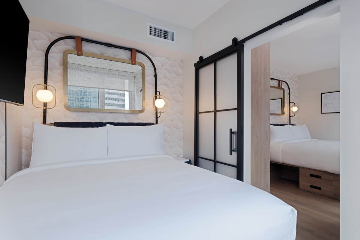 Quarto de hotel moderno com uma cama grande, espelho fixado na parede e porta deslizante para outra cama menor.