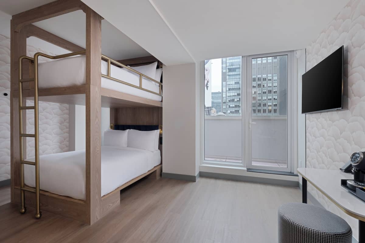Uma cama de beliche com escada, uma escrivaninha com cadeira, uma televisão e uma janela com vista para a cidade.