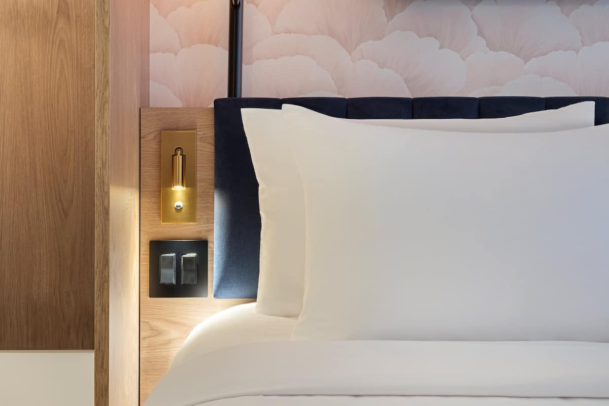 Cama bem arrumada com lençóis brancos, cabeceira escura e mesa de cabeceira de madeira com abajur e interruptor.