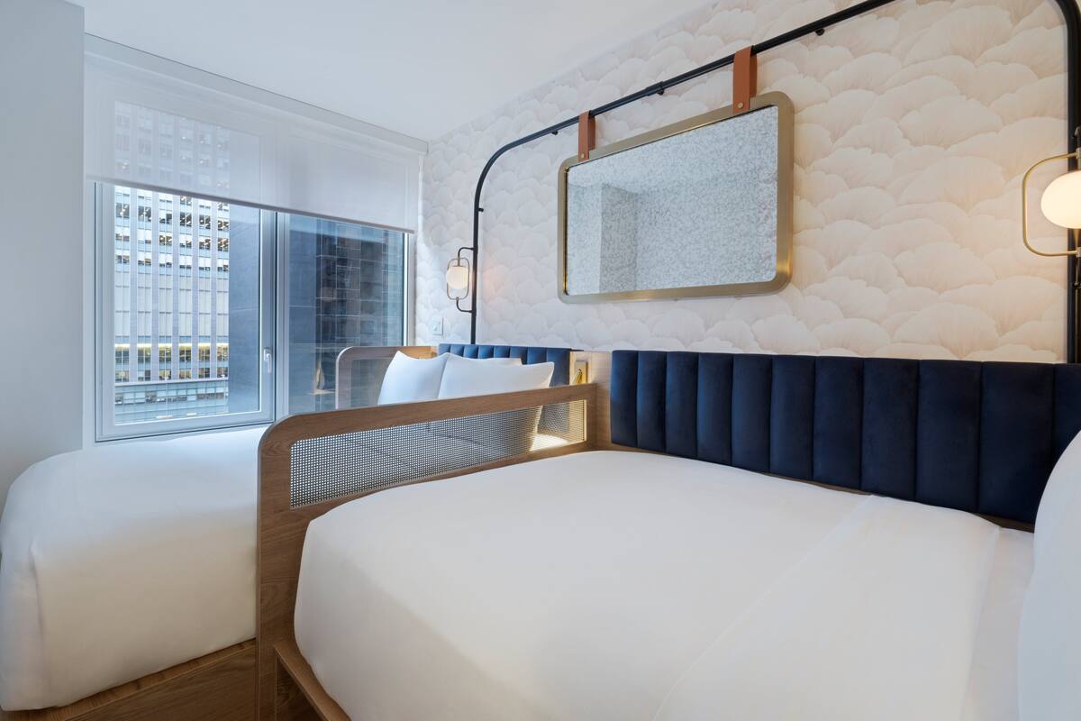 Quarto de hotel com uma cama grande, uma cama menor, uma janela vista da cidade, espelho fixado na parede e luminária de parede.