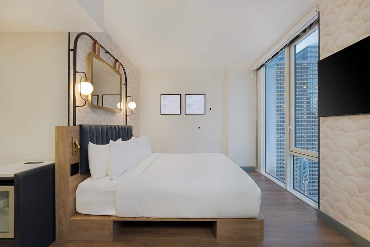 Quarto de hotel moderno com uma cama grande, uma escrivaninha, uma janela ampla com vista para a cidade e um espelho fixado na parede.