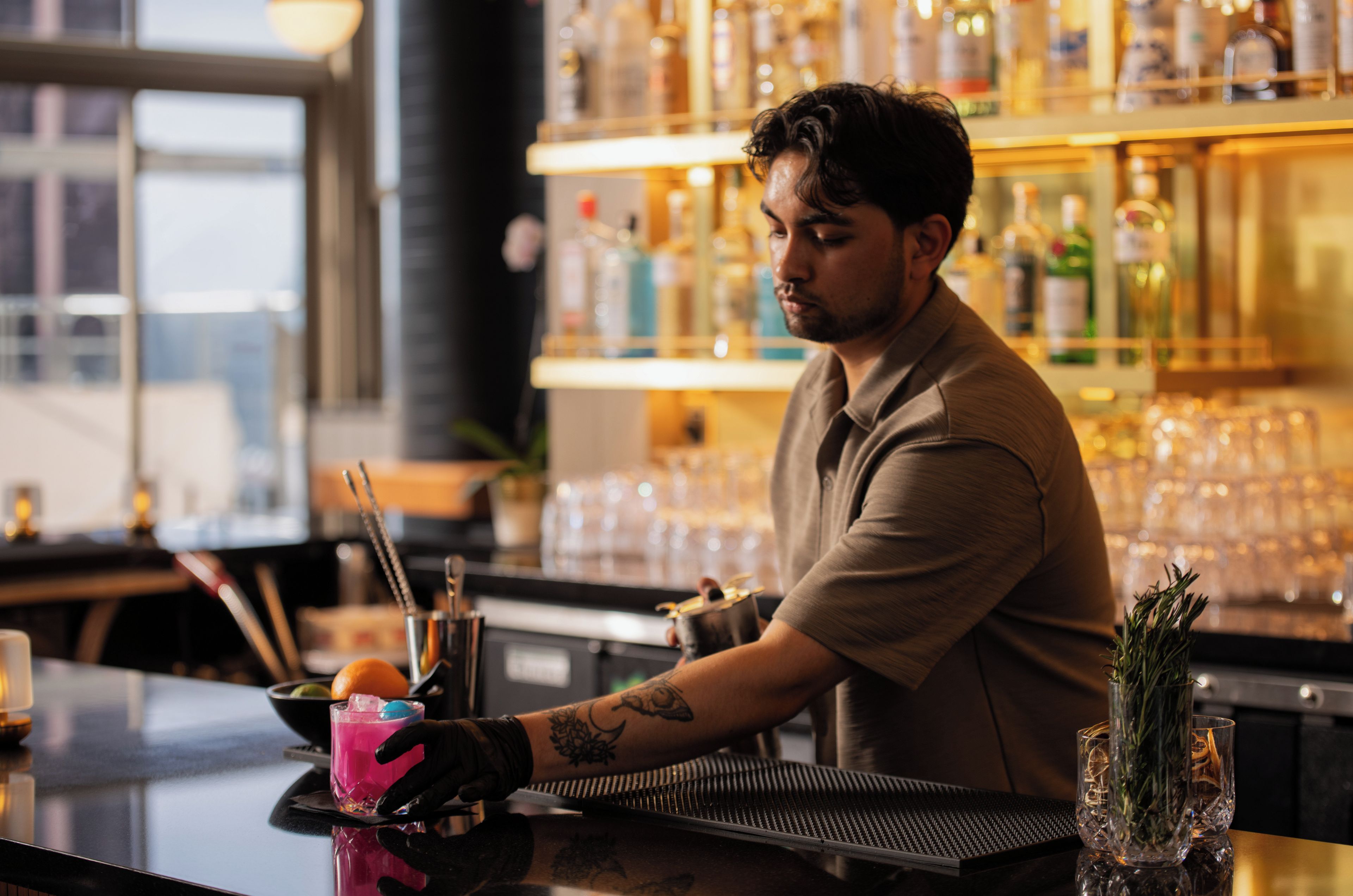 Um bartender em um bar, carregando uma bandeja com bebidas, um copo com coquetel e uma garrafa de licor ao fundo.