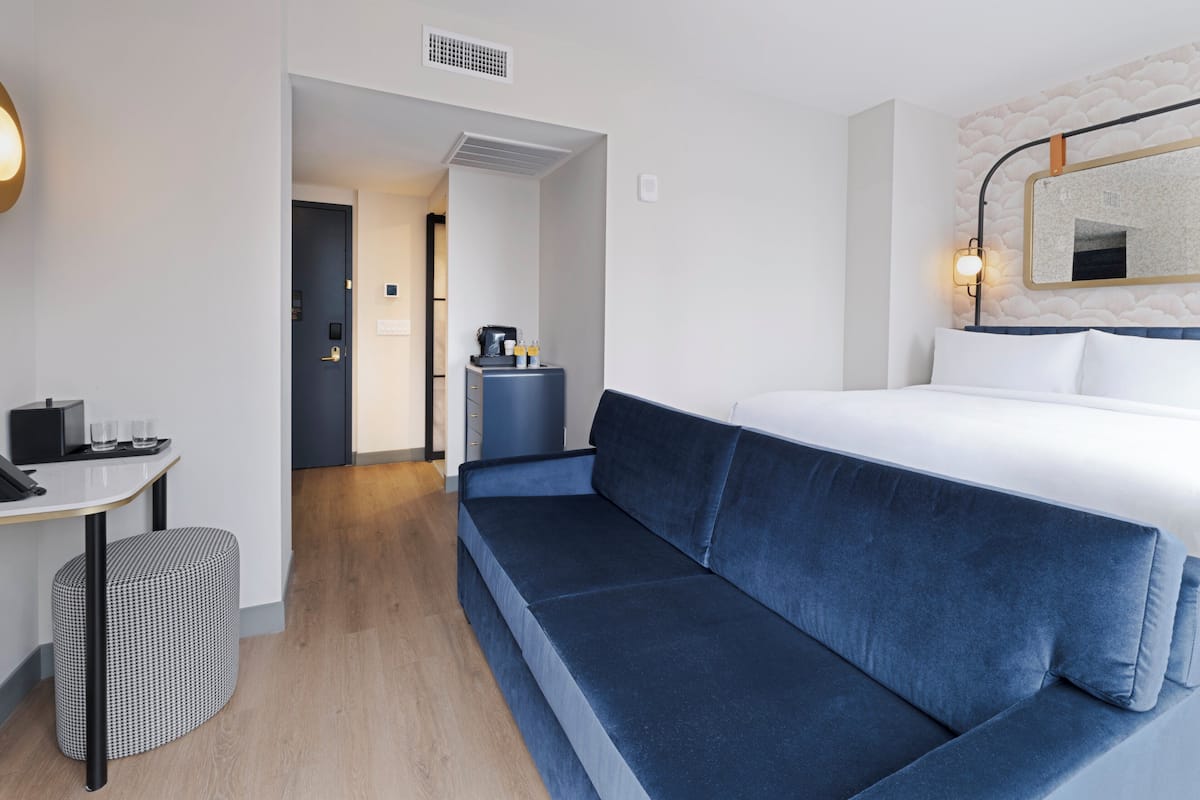 Quarto de hotel com cama, sofá, mesa pequena com abajur e escrivaninha com cadeira.