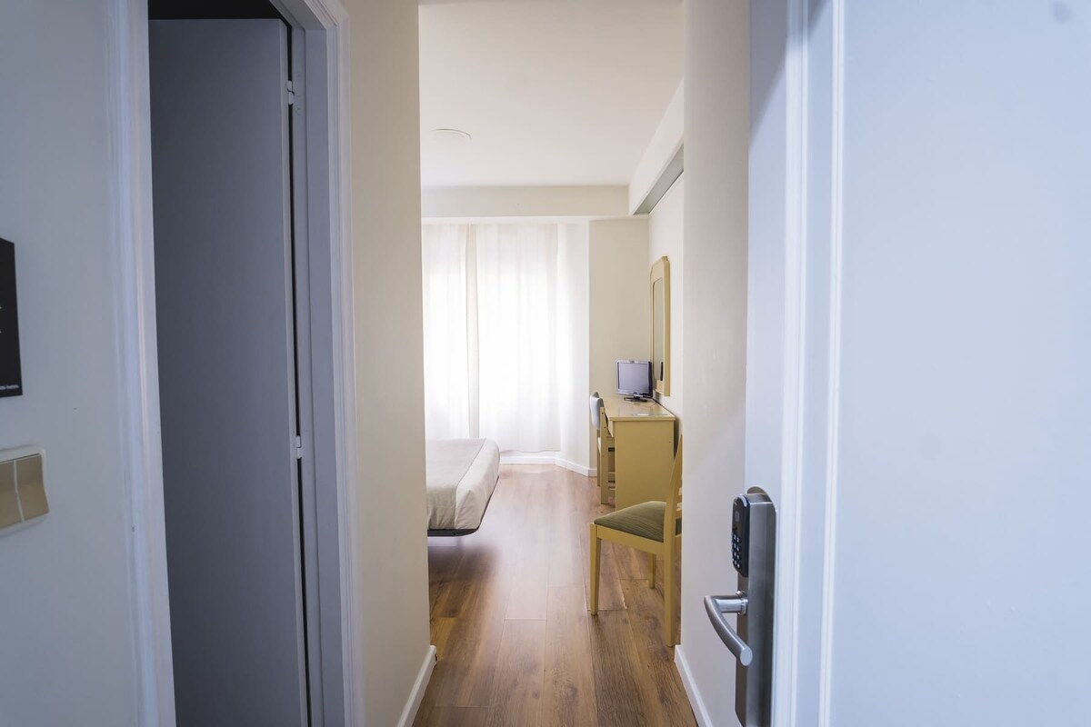 Corredor que dá para um quarto com uma cama, uma escrivaninha com um computador e uma porta com tranca de segurança.