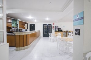 Um saguão de hotel com balcão de recepção, espaço para café e área de refeições com cadeiras e mesas brancas.