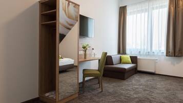 Quarto de hotel moderno com guarda-roupa de madeira, secretária com cadeira, sofá, televisão fixada na parede e uma janela grande com cortinas.