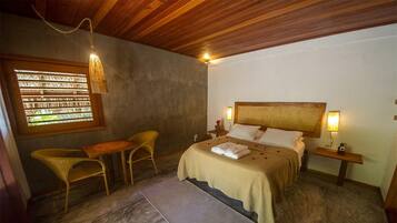 Quarto com cama, mesas de cabeceira, uma mesinha, duas cadeiras, janela com persianas e luminária pendente.