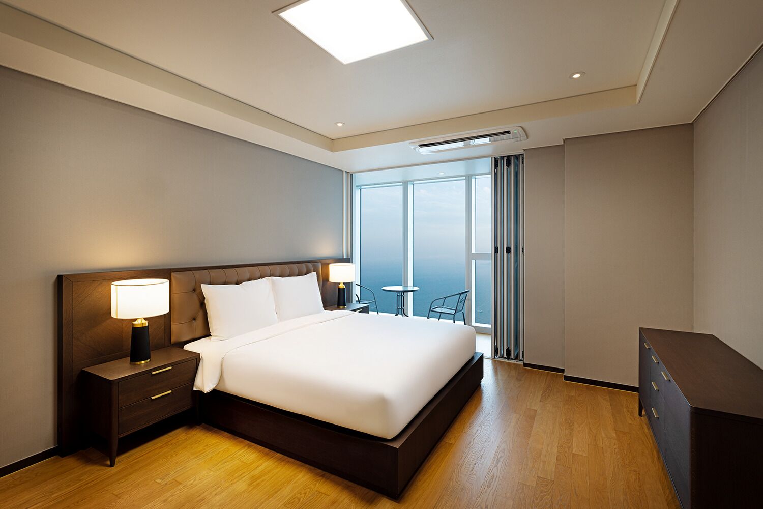 Quarto de hotel moderno com uma cama grande, duas mesas de cabeceira com luminárias, vista para o mar através de grandes janelas e uma varanda.