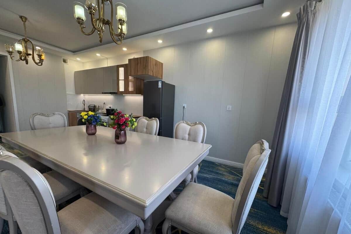 Uma sala de jantar de hotel com uma grande mesa retangular, seis cadeiras estofadas, um candelabro, uma área de cozinha e um chão coberto por carpete azul.