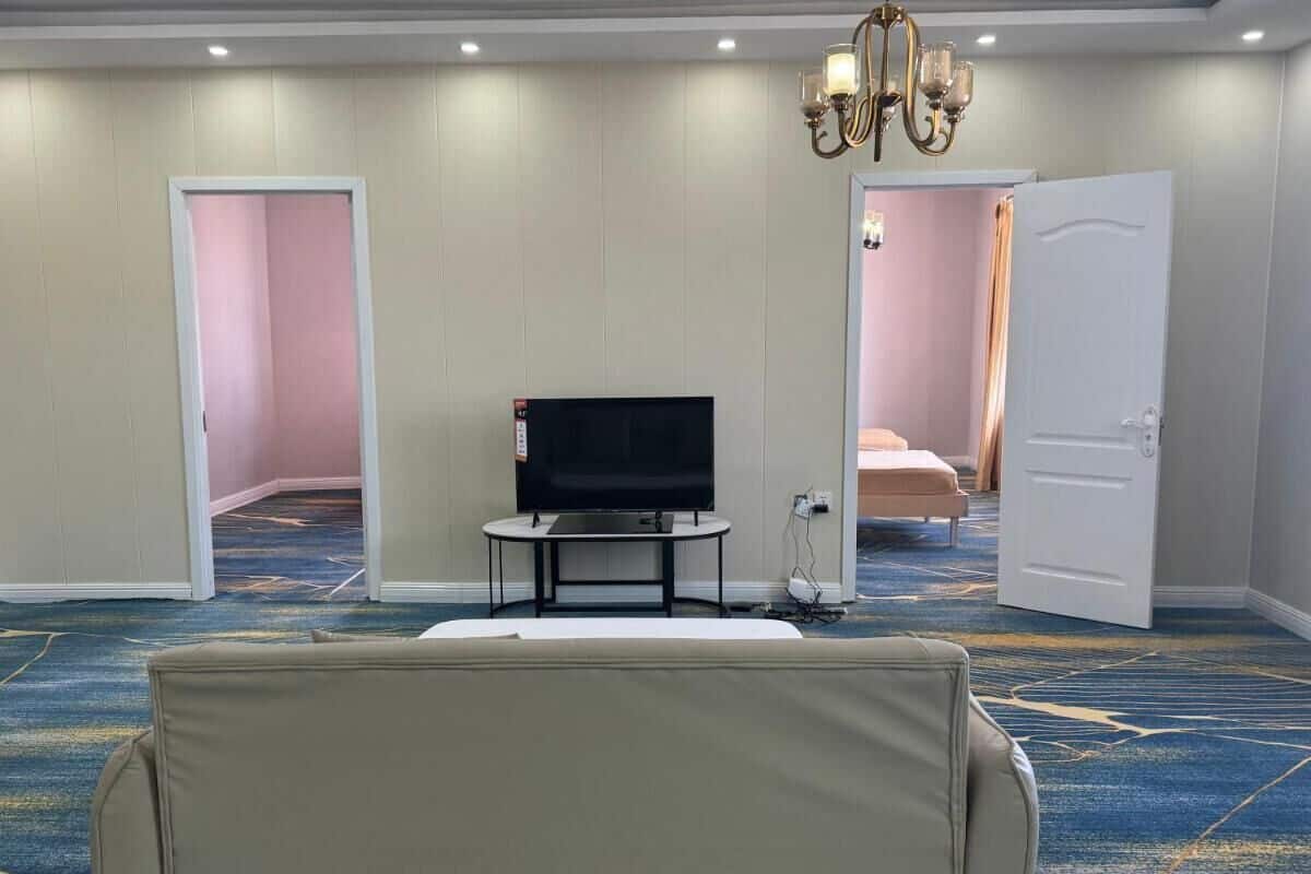 Quarto de hotel com uma grande televisão de tela plana em um suporte, um sofá bege e um tapete estampado.