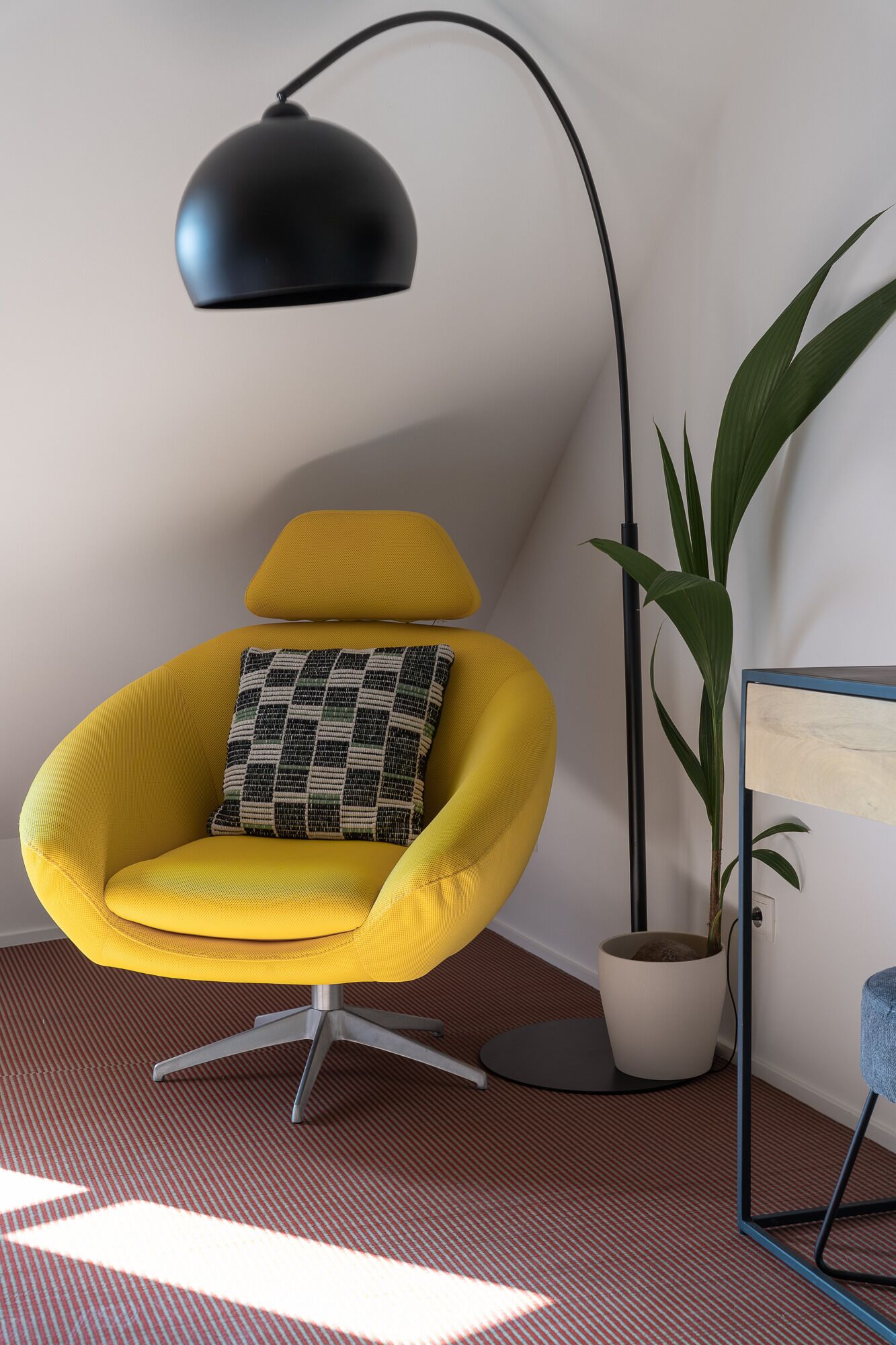 Uma poltrona amarela com uma almofada quadriculada, um abajur preto, uma planta verde alta e uma cadeira azul em um quarto com carpete listrado.