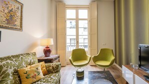 Uma sala de estar com sofá, duas poltronas, uma mesa de centro, abajur, quadro e janela com vista para prédios.