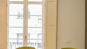 Quarto com duas cadeiras verdes, uma mesa com tampo decorativo e uma janela grande com vista para edifícios.