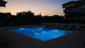 Piscina retangular com iluminação azul, rodeada por espreguiçadeiras e um deck revestido de azulejos.