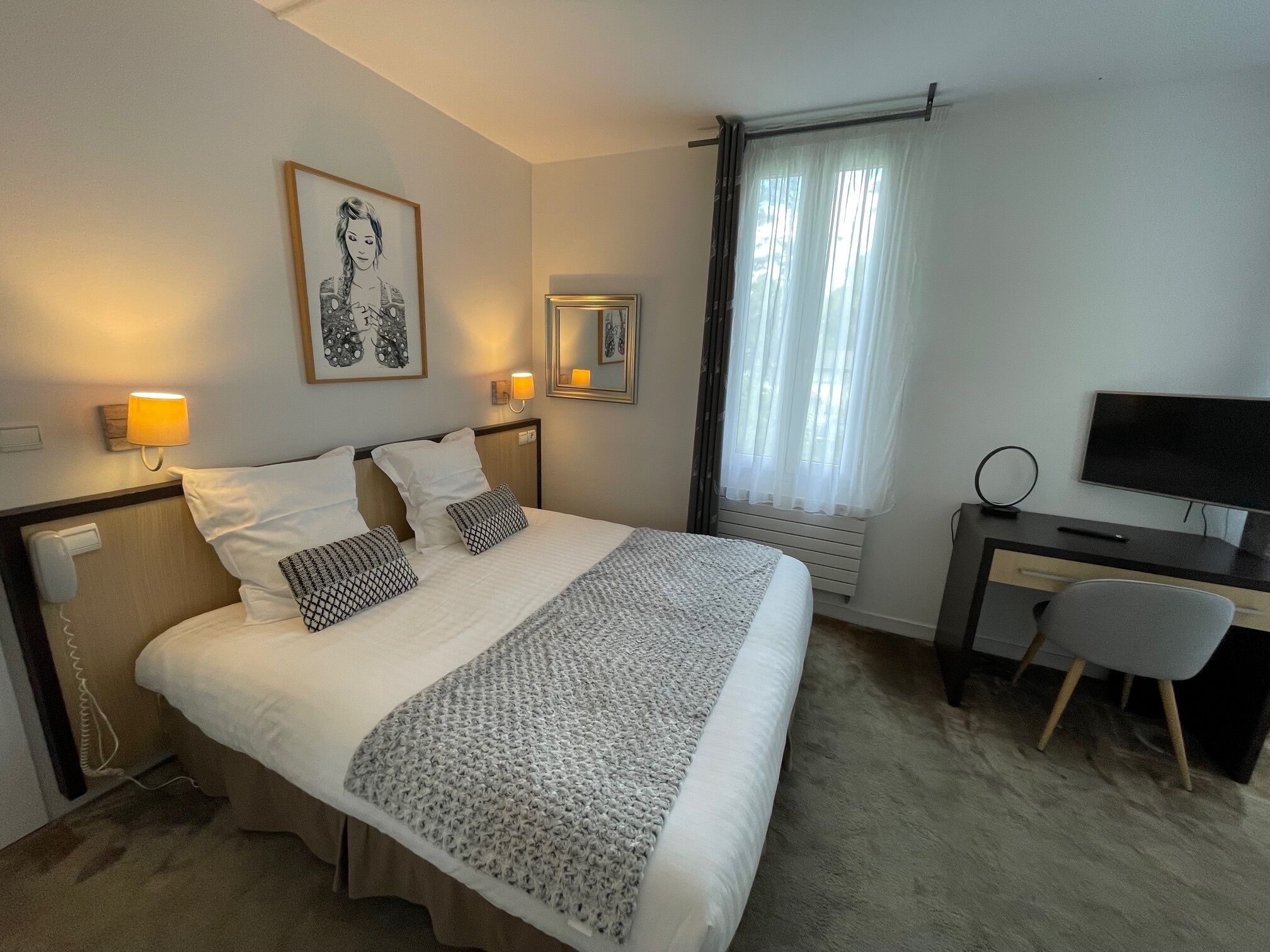 Quarto de hotel com cama, luminárias de cabeceira, escrivaninha com cadeira, televisão e janela com cortinas.