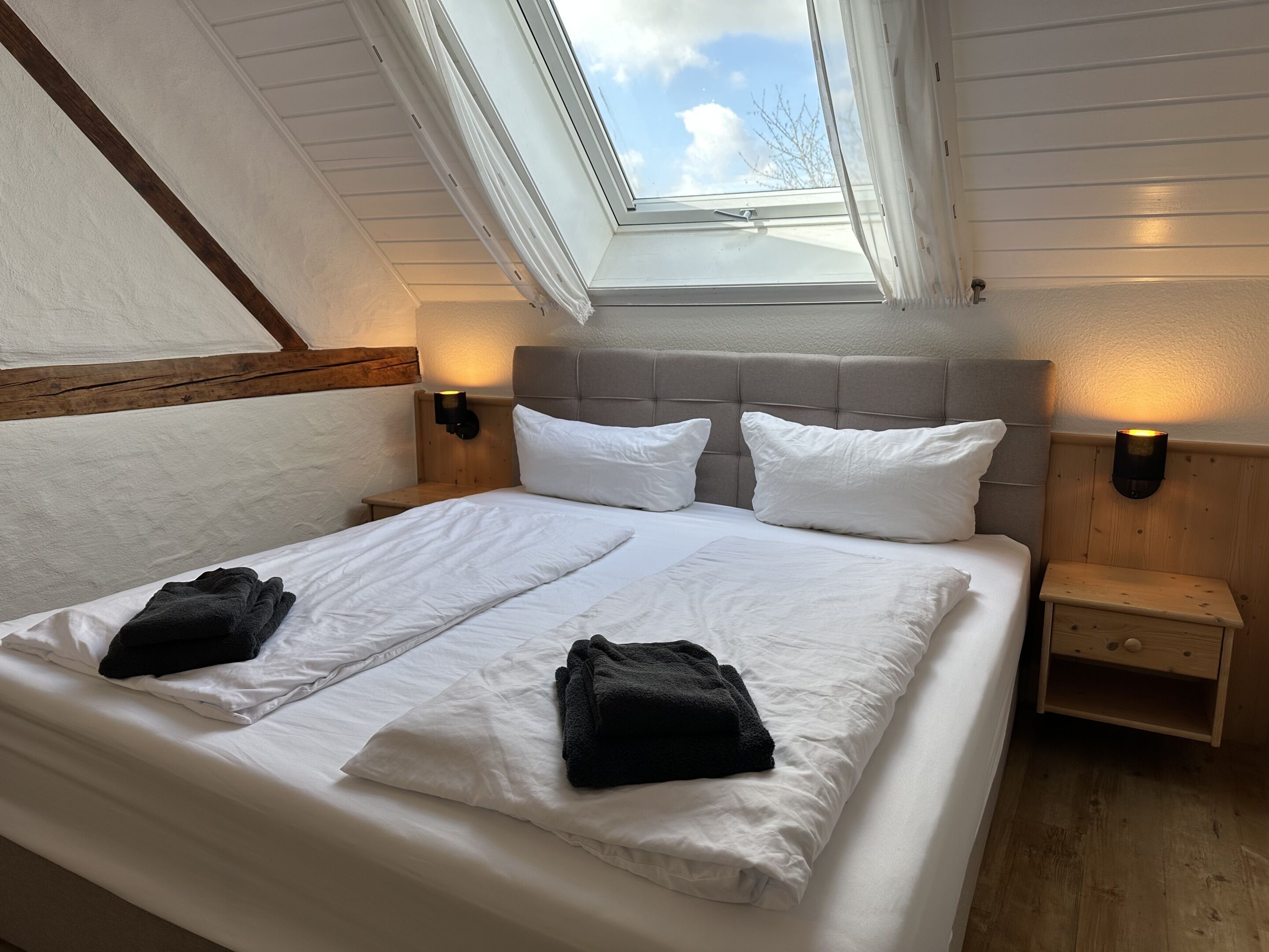 Quarto com claraboia, cama com roupa de cama branca, duas almofadas, duas toalhas pretas, cabeceira de madeira e mesas de cabeceira com luminárias fixadas na parede.