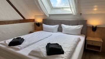 Quarto com claraboia, cama com roupa de cama branca, duas almofadas, duas toalhas pretas, cabeceira de madeira e mesas de cabeceira com luminárias fixadas na parede.