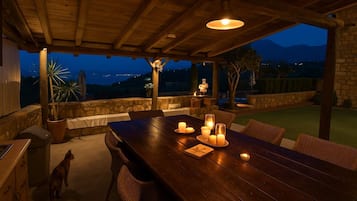 Uma mesa de jantar de madeira com velas, cadeiras de vime e vista para as montanhas ao entardecer.