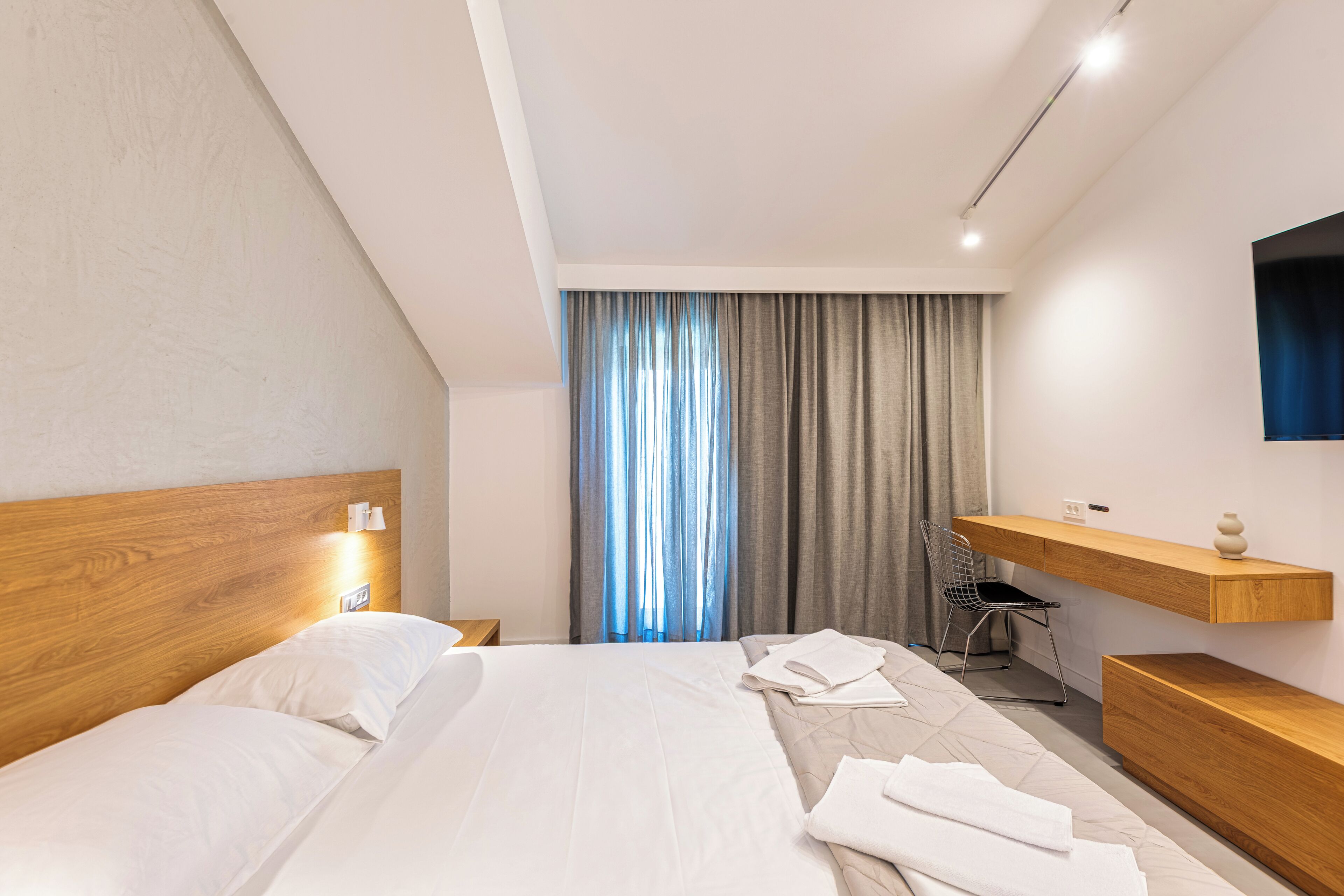 Quarto de hotel moderno com cabeceira de madeira, cama grande, escrivaninha com cadeira, televisão na parede e janela com cortinas.