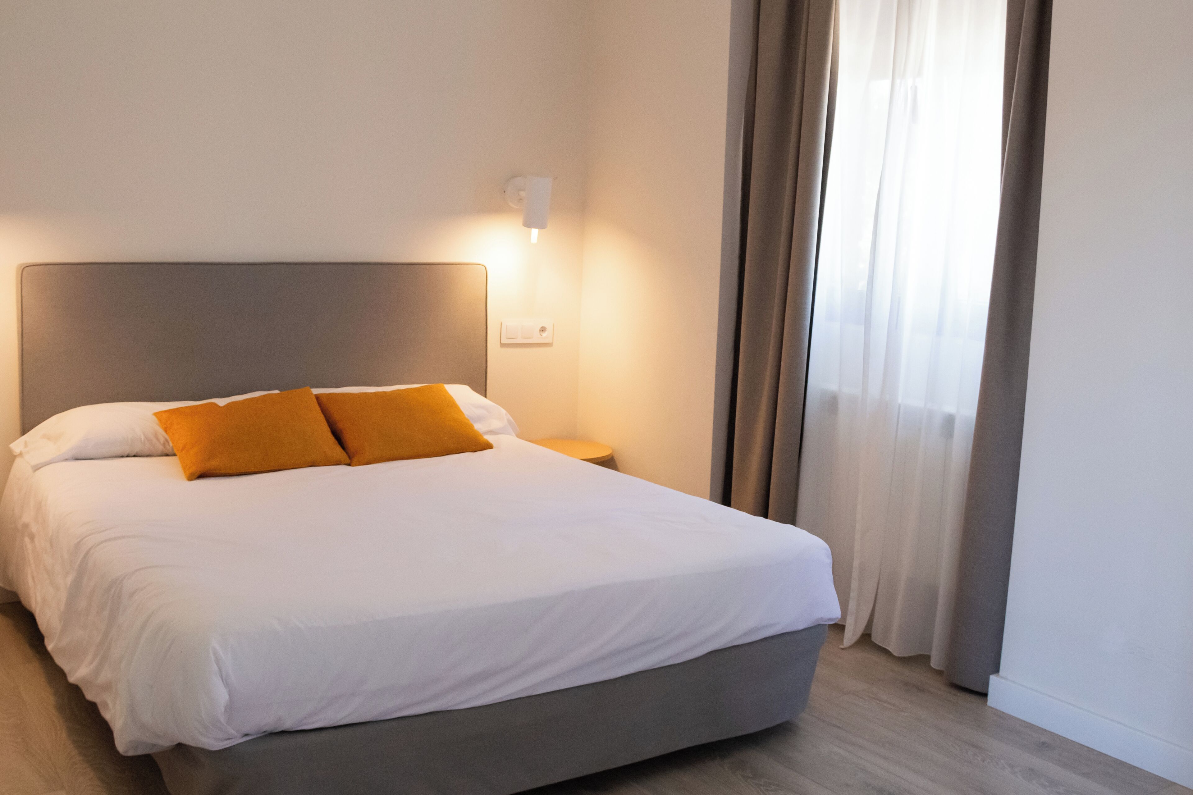 Cama bem arrumada com roupa de cama branca e laranja, luminária de cabeceira e janela com cortinas.