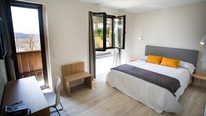 Quarto com cama, escrivaninha, cadeira e janela com vista para árvores e um edifício.