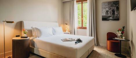 Quarto de hotel bem arrumado, com cama, mesa de cabeceira, abajur e uma pequena mesa com flores.