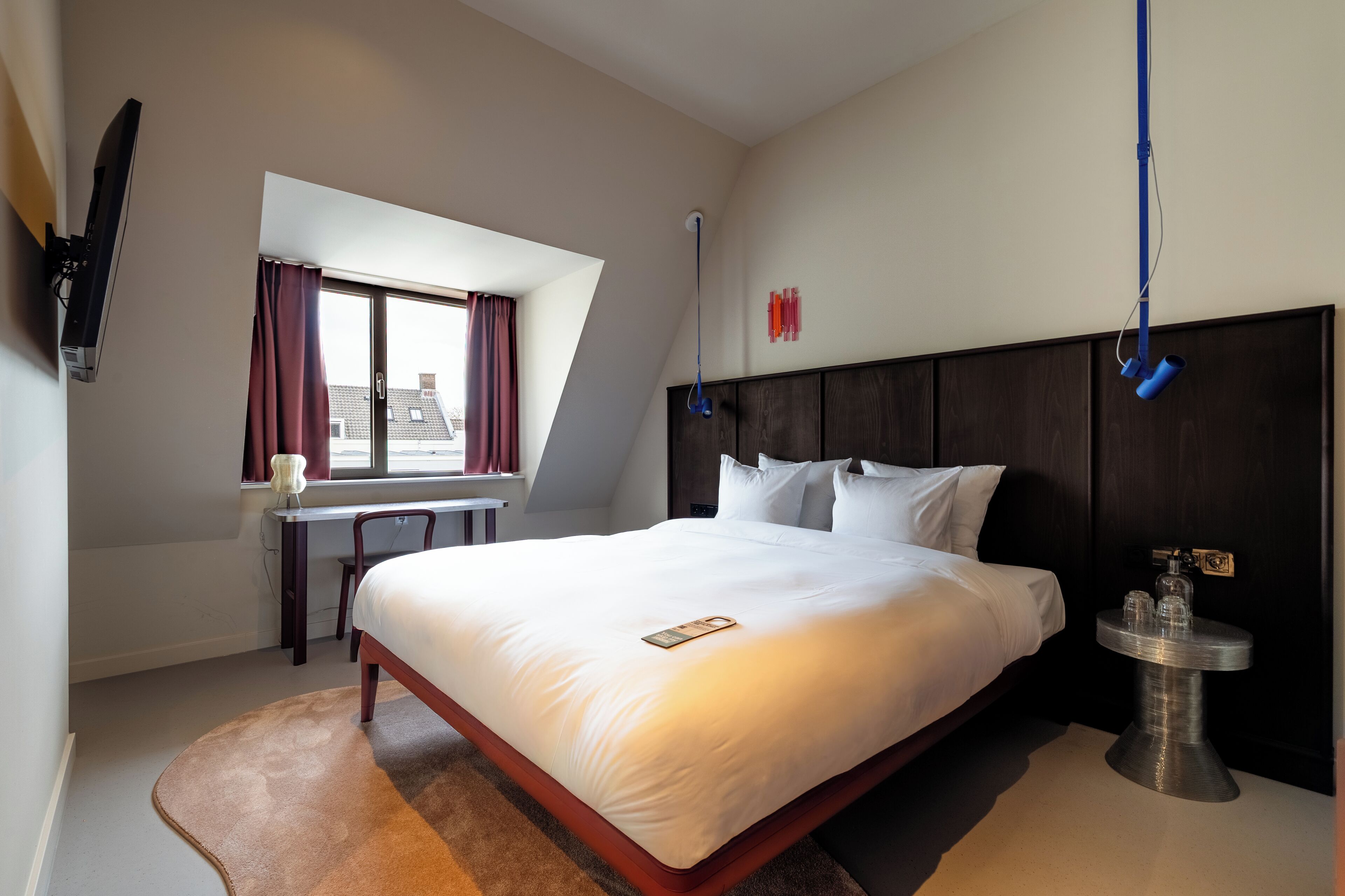Quarto de hotel com uma cama grande, uma escrivaninha com cadeira, uma janela com cortinas, uma TV na parede e uma mesinha de cabeceira com abajur.