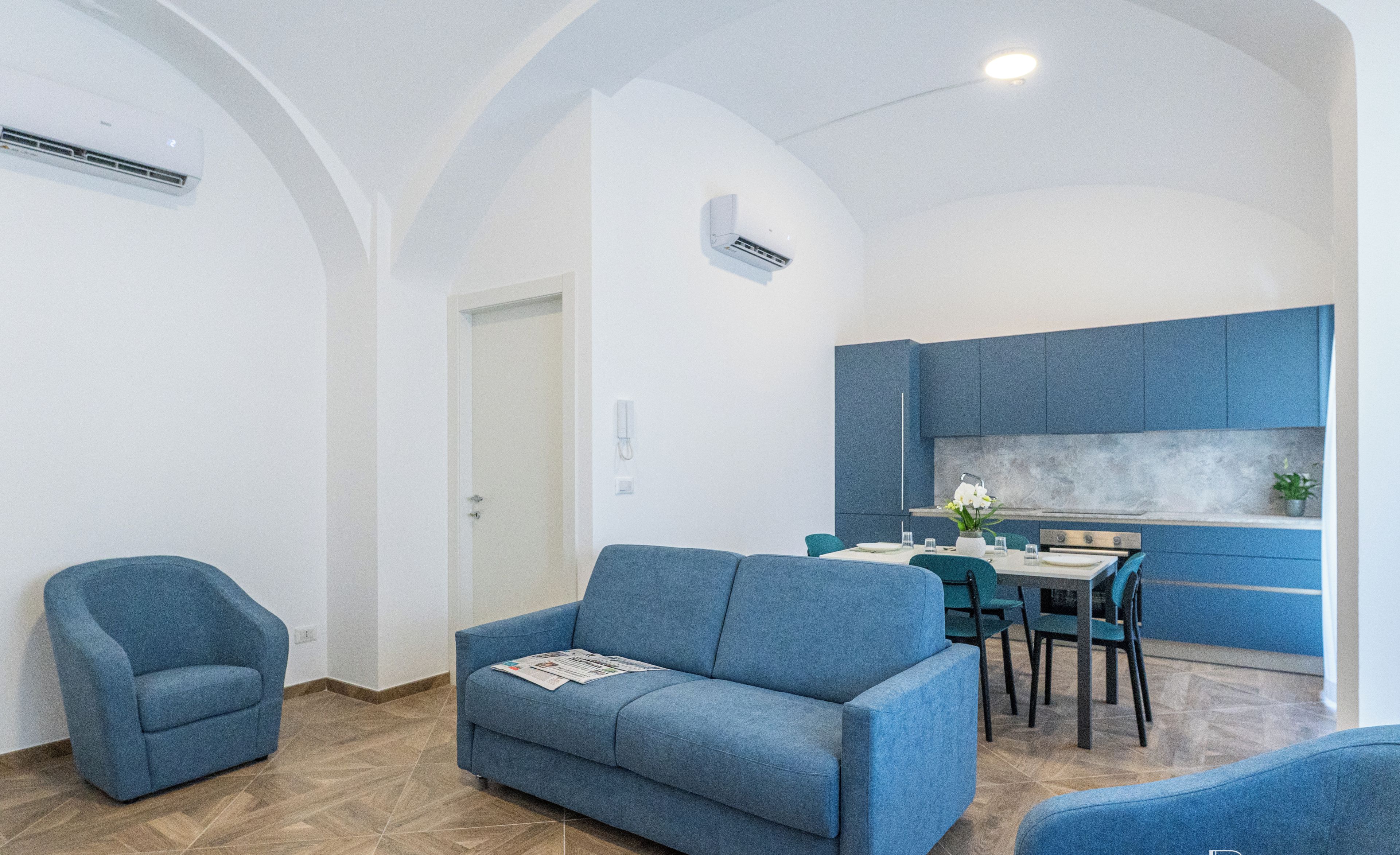 Uma sala de estar moderna com um sofá azul, uma poltrona coordenada e um espaço de refeições com mesa e cadeiras. Há uma cozinha com armários azuis e bancada.