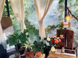 Quarto com uma grande janela, cortinas e diversas plantas. Há uma mesa de madeira com um vaso de flores e uma pequena prateleira com objetos decorativos.