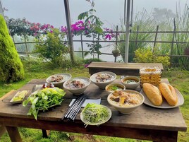 Mesa posta ao ar livre com diversos pratos, incluindo sopas em tigelas e pães em travessas, rodeada por folhagem e flores.