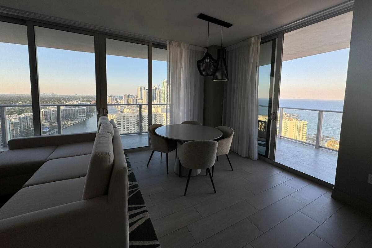 Uma sala de estar moderna com sofá, mesa de jantar e cadeiras. Possui grandes janelas com vista para a cidade e oceano.