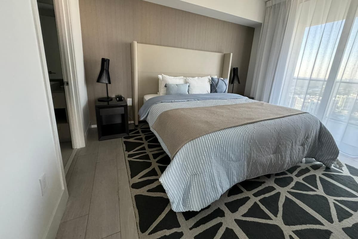 Quarto de hotel moderno com uma cama grande, mesa de cabeceira, luminária e vista para a cidade pela janela.