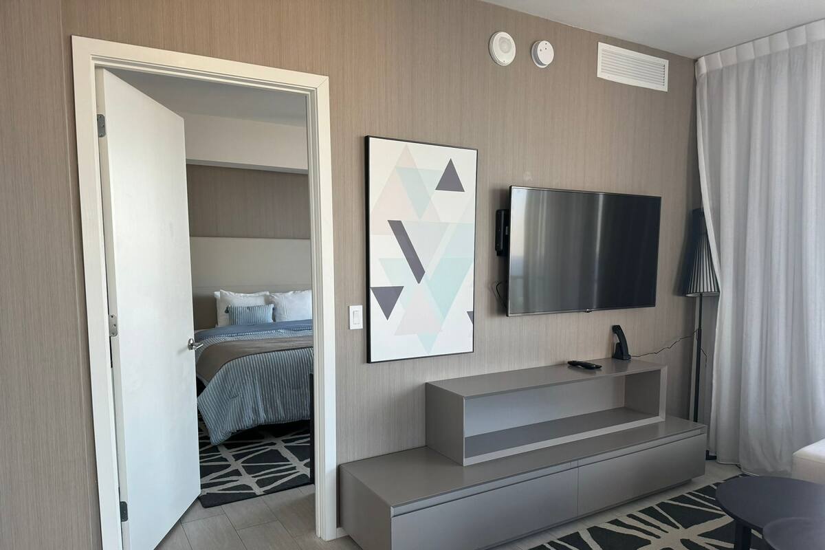 Quarto de hotel moderno com uma cama, televisão fixada na parede, um pequeno espaço de estar e uma porta que dá para outro quarto.