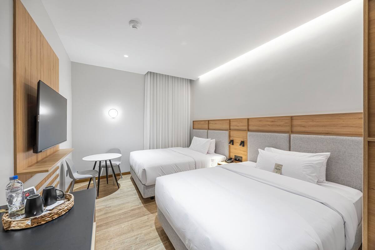 Quarto de hotel com duas camas, televisão, uma mesinha com uma cesta e uma cadeira branca.