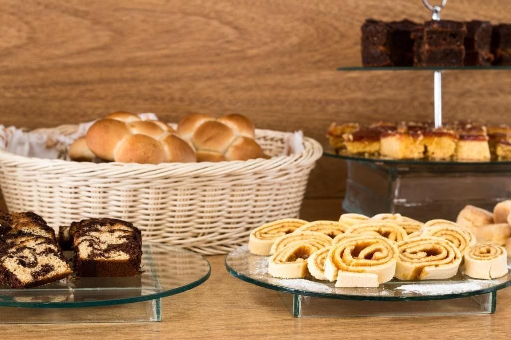 Exposição de diversos produtos de panificação, incluindo pães, um bolo e doces, em suportes de vidro.