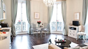 Uma sala de estar ampla, com área de jantar, um candelabro e janelas grandes com cortinas.