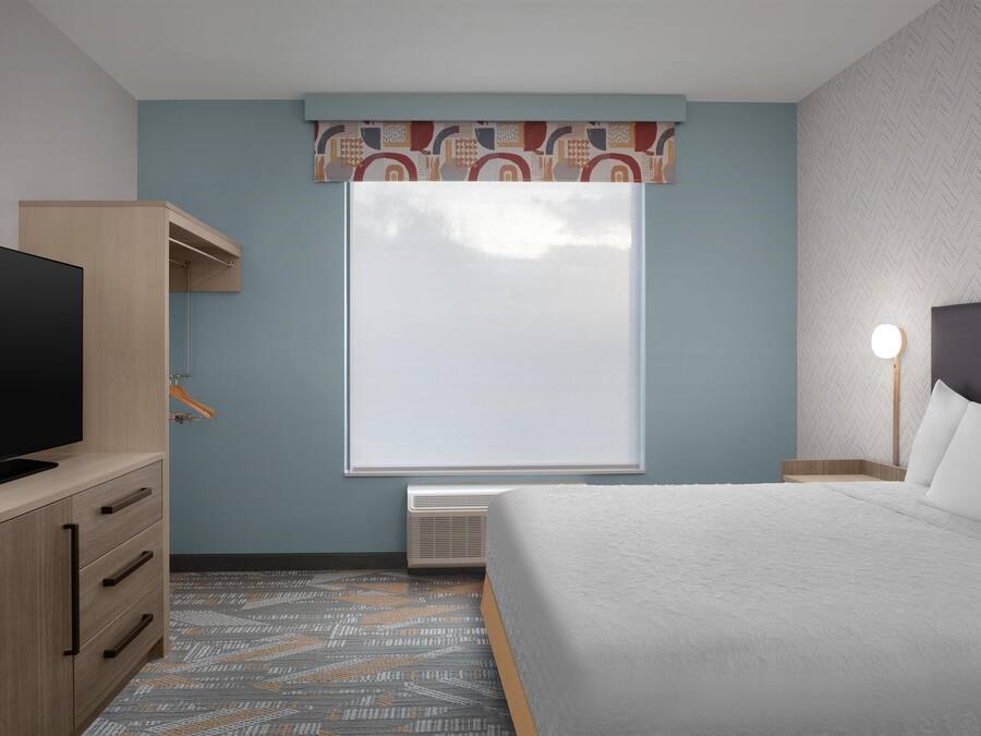 Quarto de hotel com cama, televisão, guarda-roupa, janela com cortina e mesa de cabeceira com abajur.