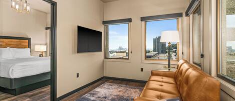 Quarto de hotel com cama, sofá, televisão e amplas janelas com vista para a cidade.
