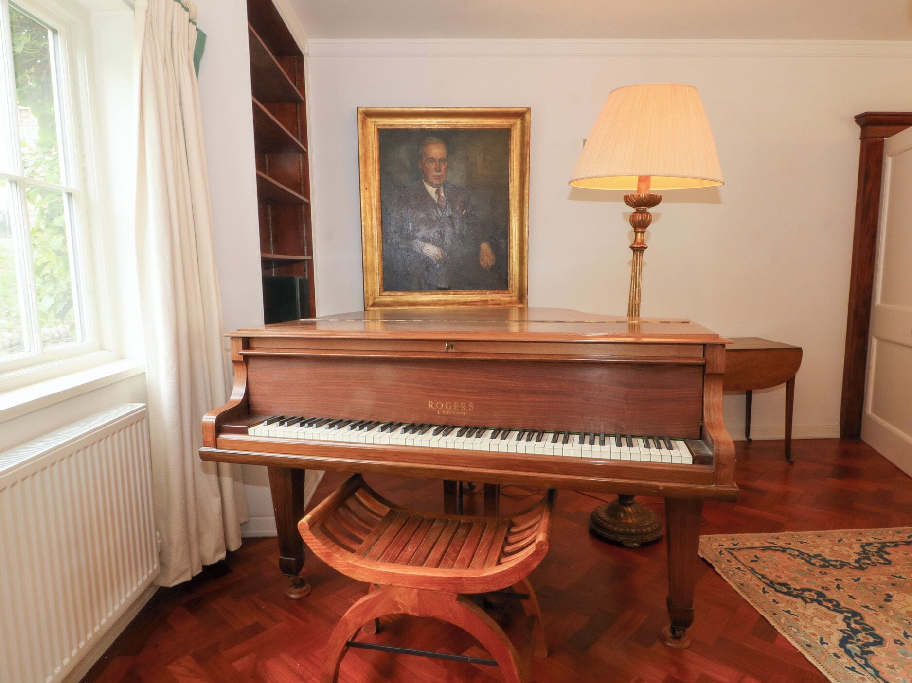 Um piano de cauda em um quarto com um radiador, uma cadeira de madeira, um abajur, um retrato emoldurado e um tapete estampado.