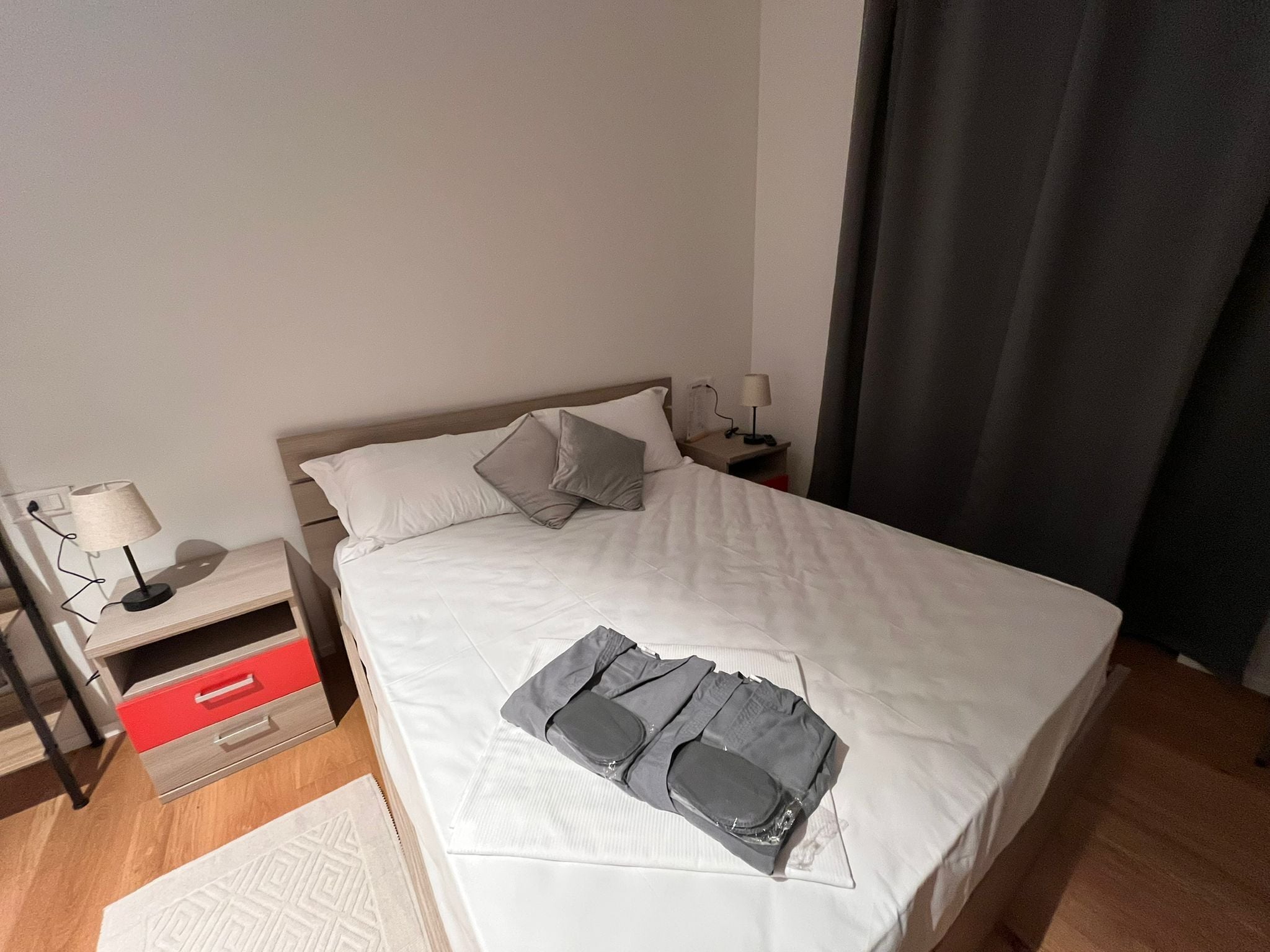 Uma cama bem arrumada com almofadas, uma mesinha de cabeceira com luminárias e um móvel com gavetas vermelho.