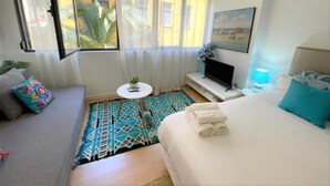Quarto de hotel com cama, televisão, pequena mesa com planta, sofá e janela com cortinas.