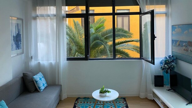 Uma sala de estar com sofá, mesa de centro com planta, televisão e uma grande janela com vista para palmeiras e um edifício amarelo.
