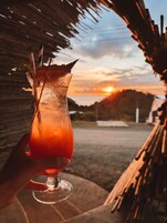 Uma mão segurando um copo com bebida vermelha e canudo, com o céu do pôr do sol ao fundo.
