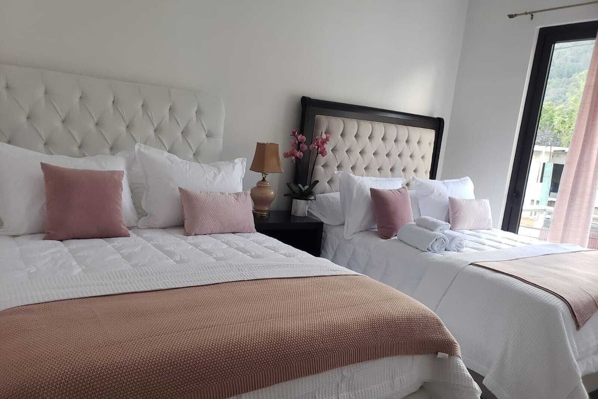 Quarto de hotel com duas camas, roupa de cama branca, cabeceira estofada, mesa de cabeceira com abajur e janela com vista.