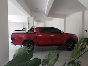 Uma caminhonete vermelha estacionada em uma garagem de paredes brancas, com um ventilador de teto e um vaso de planta.