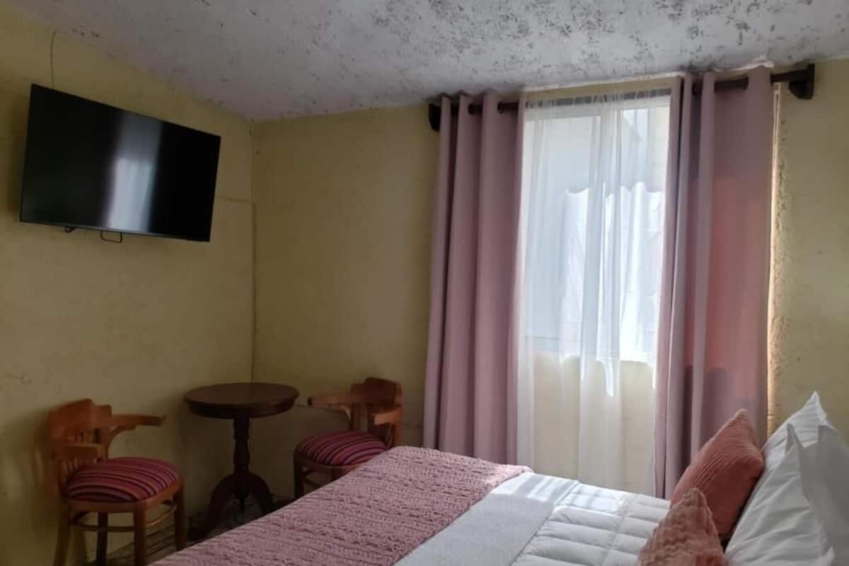 Quarto com cama, televisão na parede, mesa pequena, duas cadeiras e janela com cortinas.