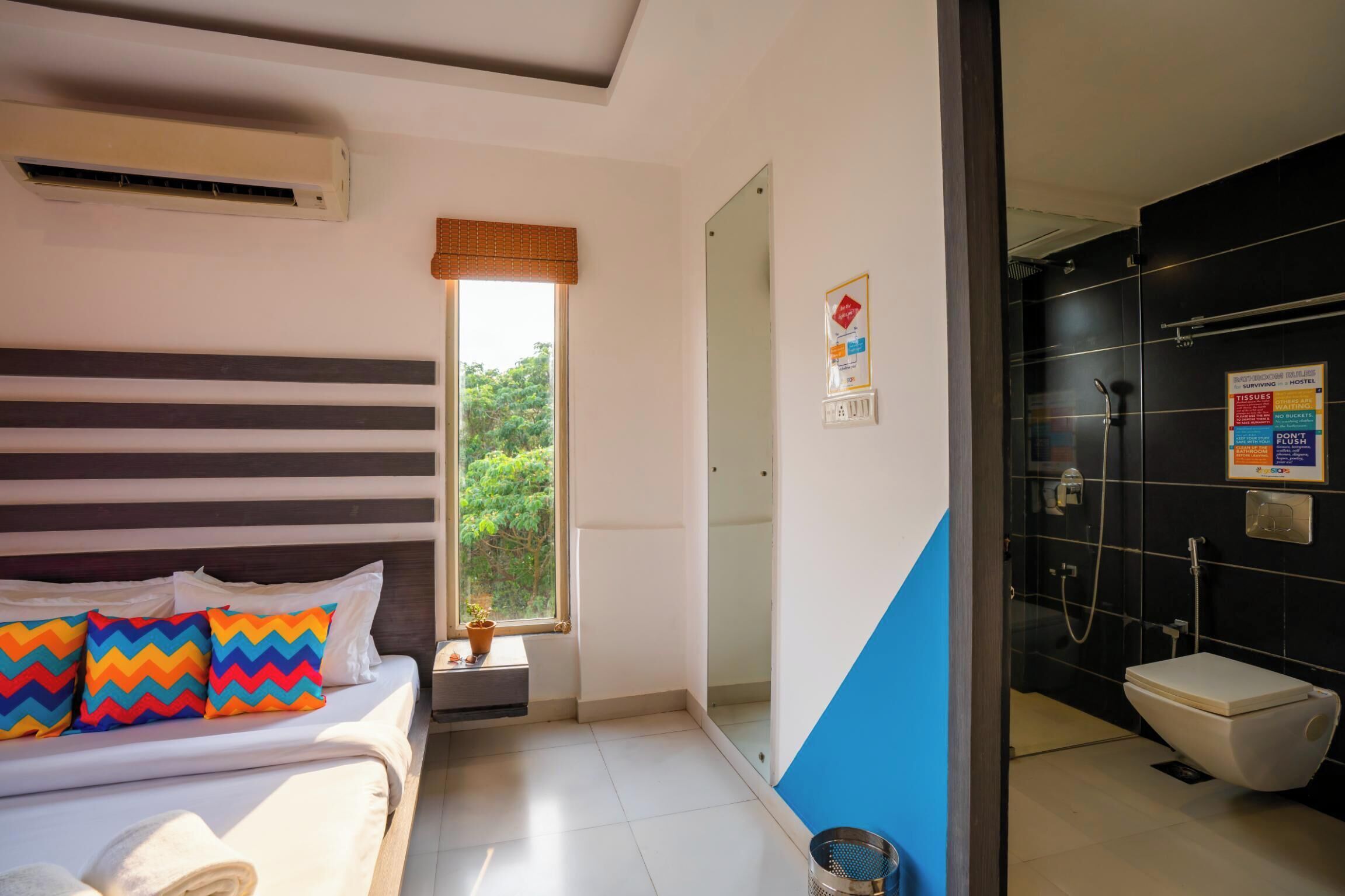 Quarto de hotel com cama, janela vista para o verde, banheiro com chuveiro e vaso sanitário, e parede com um aviso.