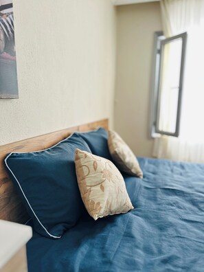 Uma cama com roupa de cama azul e duas almofadas, uma delas com padrão floral, em um quarto com janela e um quadro na parede.