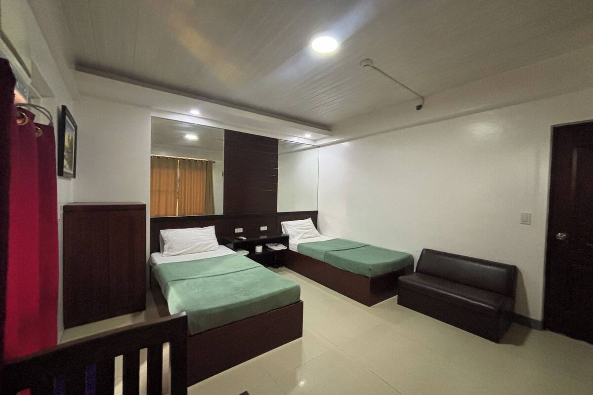 Quarto de hotel com duas camas de solteiro, um sofá, um guarda-roupa de madeira, um espelho e uma porta.