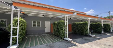 Área de estacionamento coberta com garagem, porta e pequeno jardim com tapete verde e arbustos.